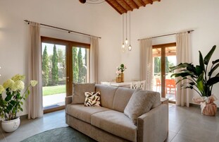 Sala de estar com sofá bege, mesa de centro e uma grande planta em vaso. Há cortinas nas janelas e vista para o exterior.