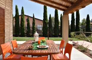 Uma mesa de madeira com cadeiras laranja sob uma pérgula, em um jardim com ciprestes altos e um edifício ao fundo.