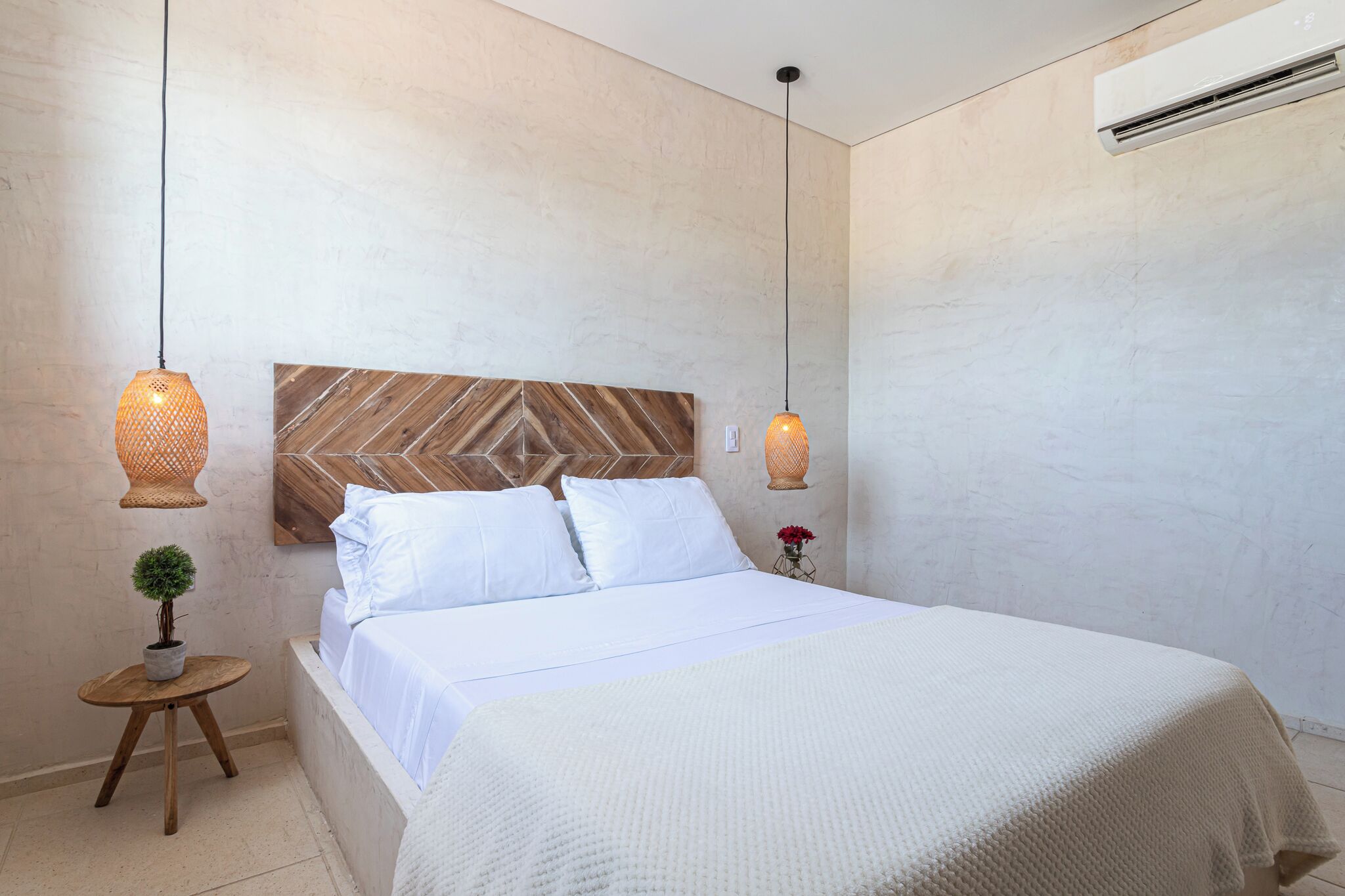Quarto com cama, duas luminárias pendentes, uma mesinha com planta e ar-condicionado fixado na parede.