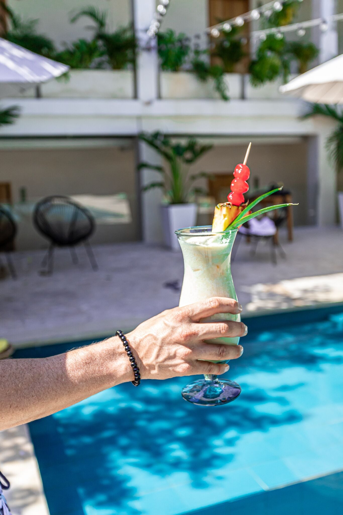 Uma mão segurando um drink com um espeto de abacaxi, perto de uma piscina com área de estar externa e plantas.
