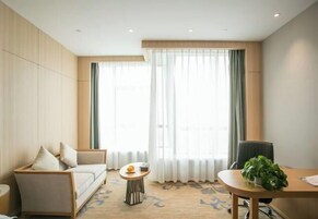 Quarto de hotel moderno com sofá, uma mesinha redonda com uma cesta de frutas, uma cadeira e um vaso de plantas.