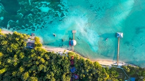 Vista aérea de uma praia tropical com águas turquesa cristalinas, um píer de madeira e um pequeno barco.
