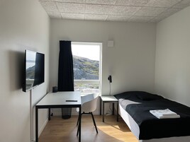 Quarto com cama, escrivaninha, cadeira, televisão e janela com vista para uma paisagem rochosa.