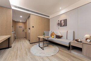 Quarto de hotel moderno com piso de madeira, tapete redondo, sofá, mesa pequena e mesa de cabeceira com abajur.