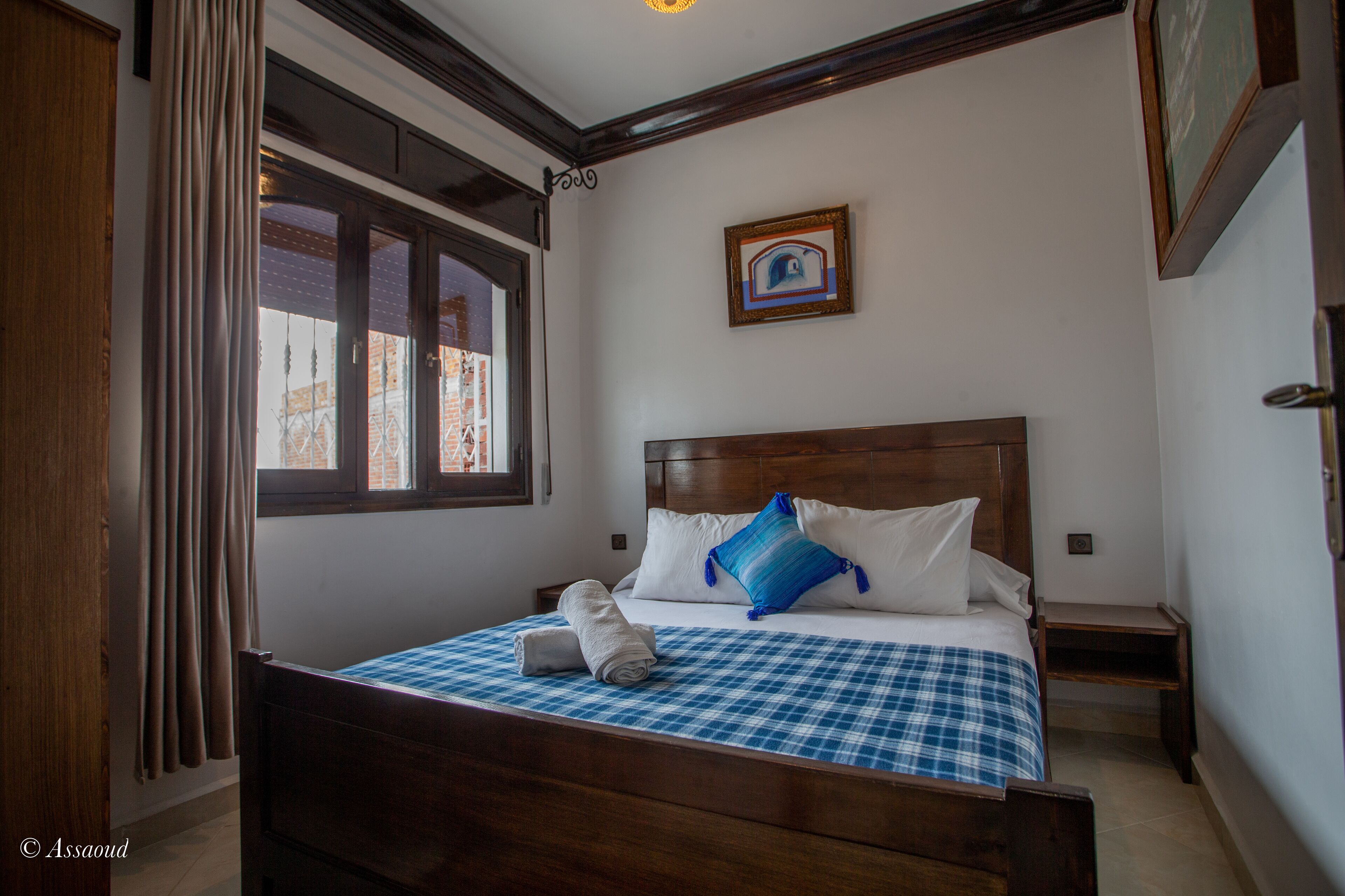 Cama bem arrumada com colcha xadrez, um travesseiro azul e um travesseiro branco. Tem cabeceira de madeira, uma mesinha de cabeceira pequena e um quadro na parede.