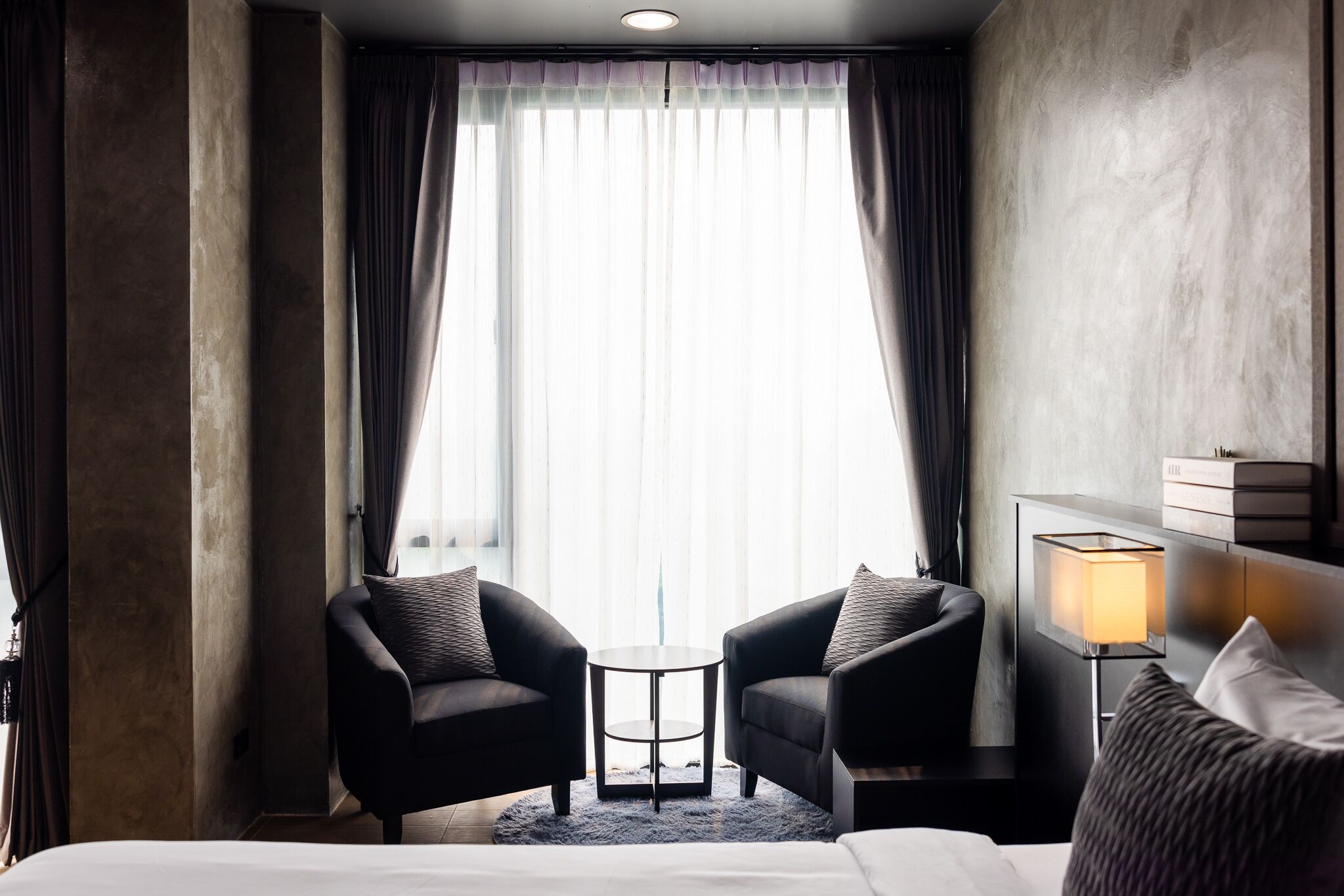 Quarto de hotel com cama, duas poltronas, uma mesa pequena, abajur e vista através de cortinas leves.