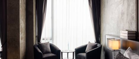 Quarto de hotel com cama, duas poltronas, uma mesa pequena, abajur e vista através de cortinas leves.
