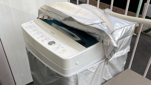 Máquina de lavar roupa pequena e branca com painel de controle, capa protetora e um pequeno armário com garrafas.