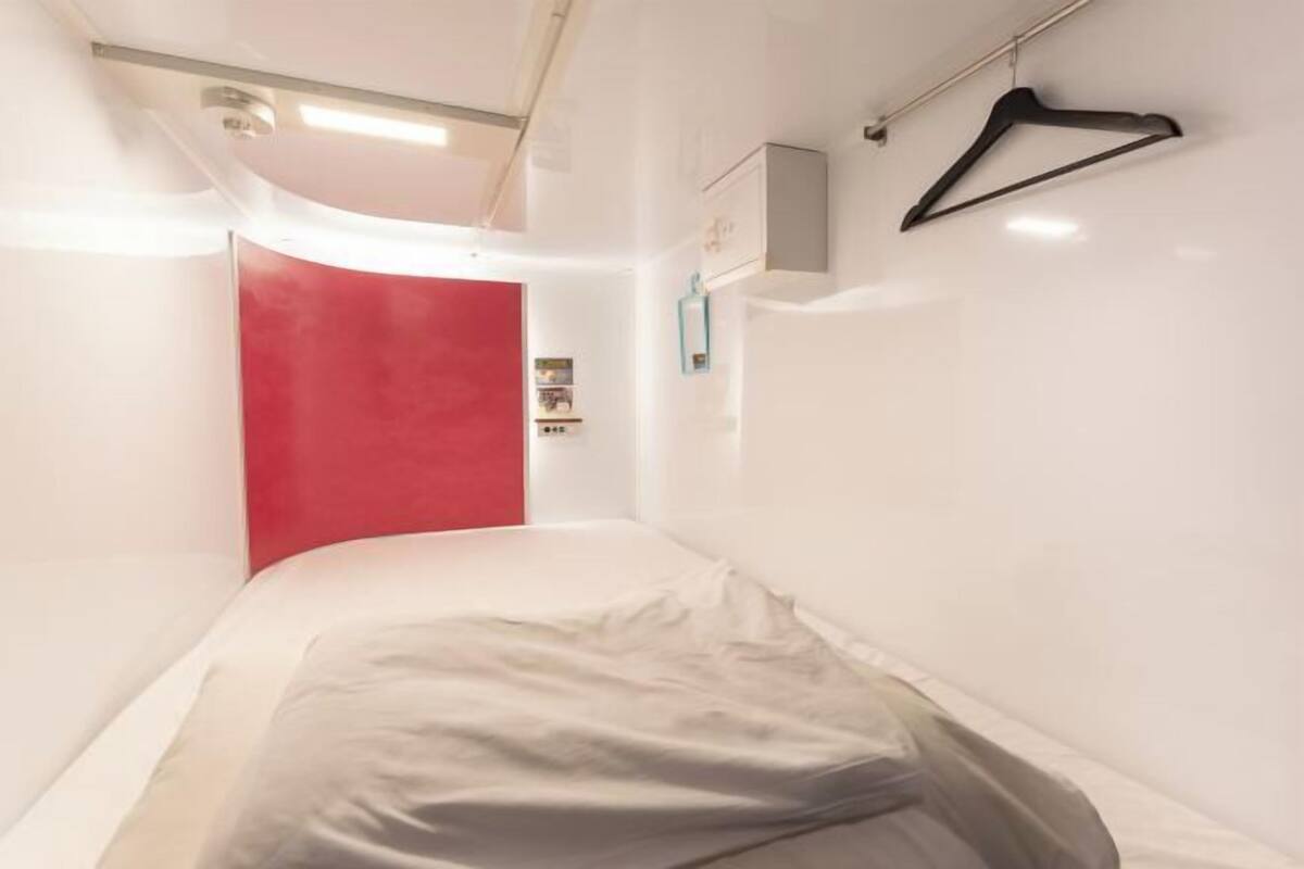 Quarto de hotel estreito com uma cama, uma parede de destaque vermelha, um cabide preto e uma pequena prateleira com objetos.