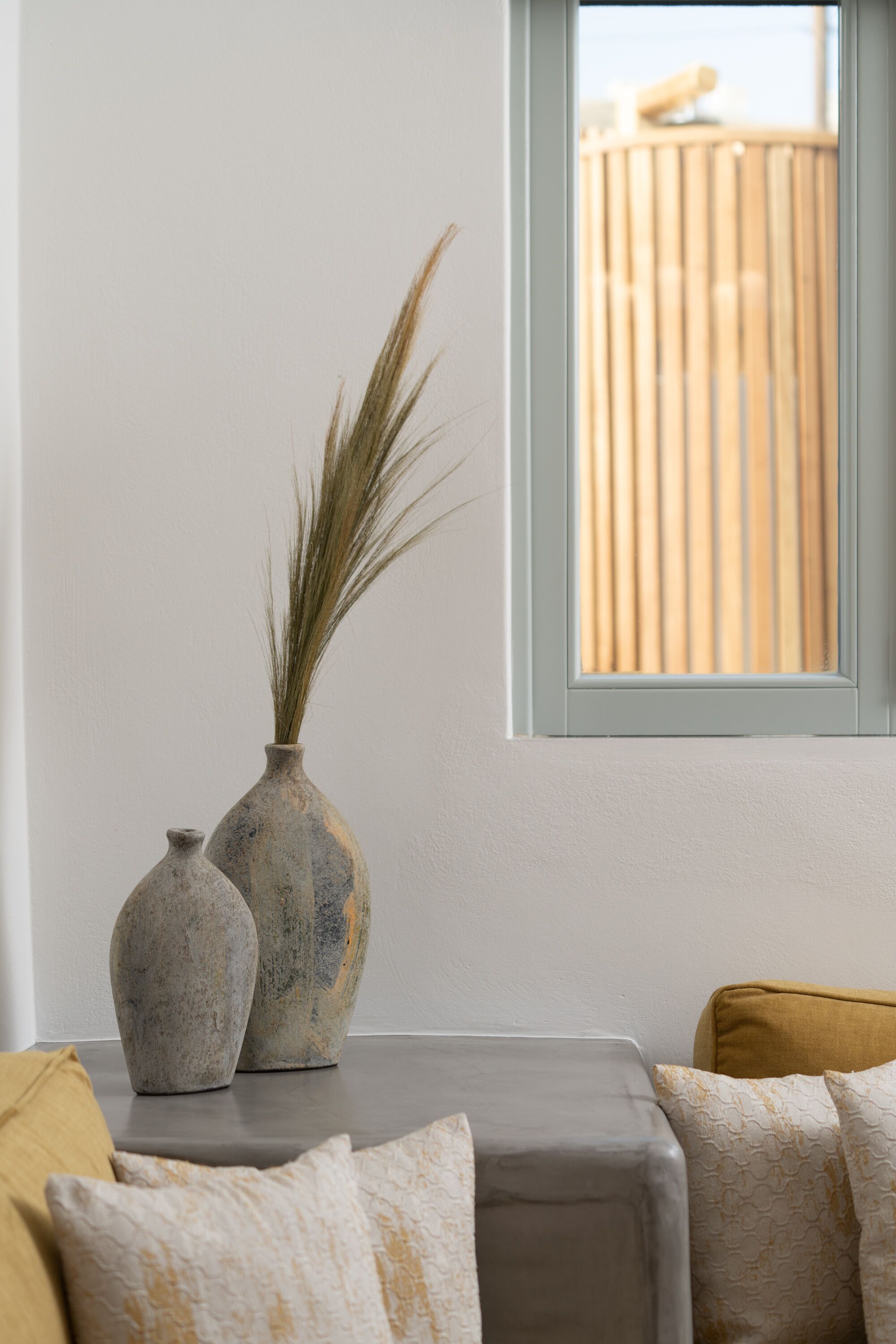 Quarto com sofá cinzento, dois vasos com plantas secas e uma janela com persianas.
