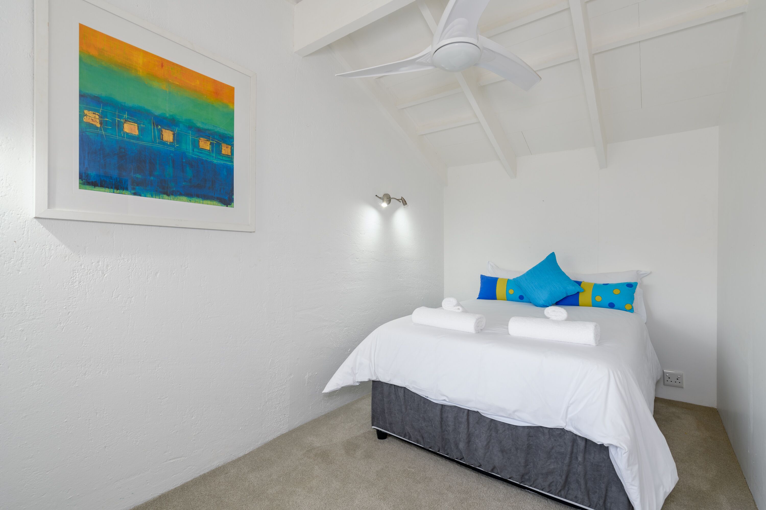 Quarto com cama, paredes brancas, ventilador de teto e um quadro na parede.
