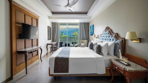 Quarto de hotel com uma cama grande, um ventilador de teto, uma televisão, varanda com vista e uma escrivaninha com abajur.
