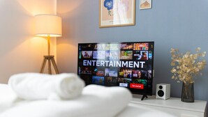 Quarto de hotel com televisão de tela plana exibindo diversos canais de entretenimento, uma luminária, um alto-falante e um vaso com flores secas na mesinha de cabeceira.