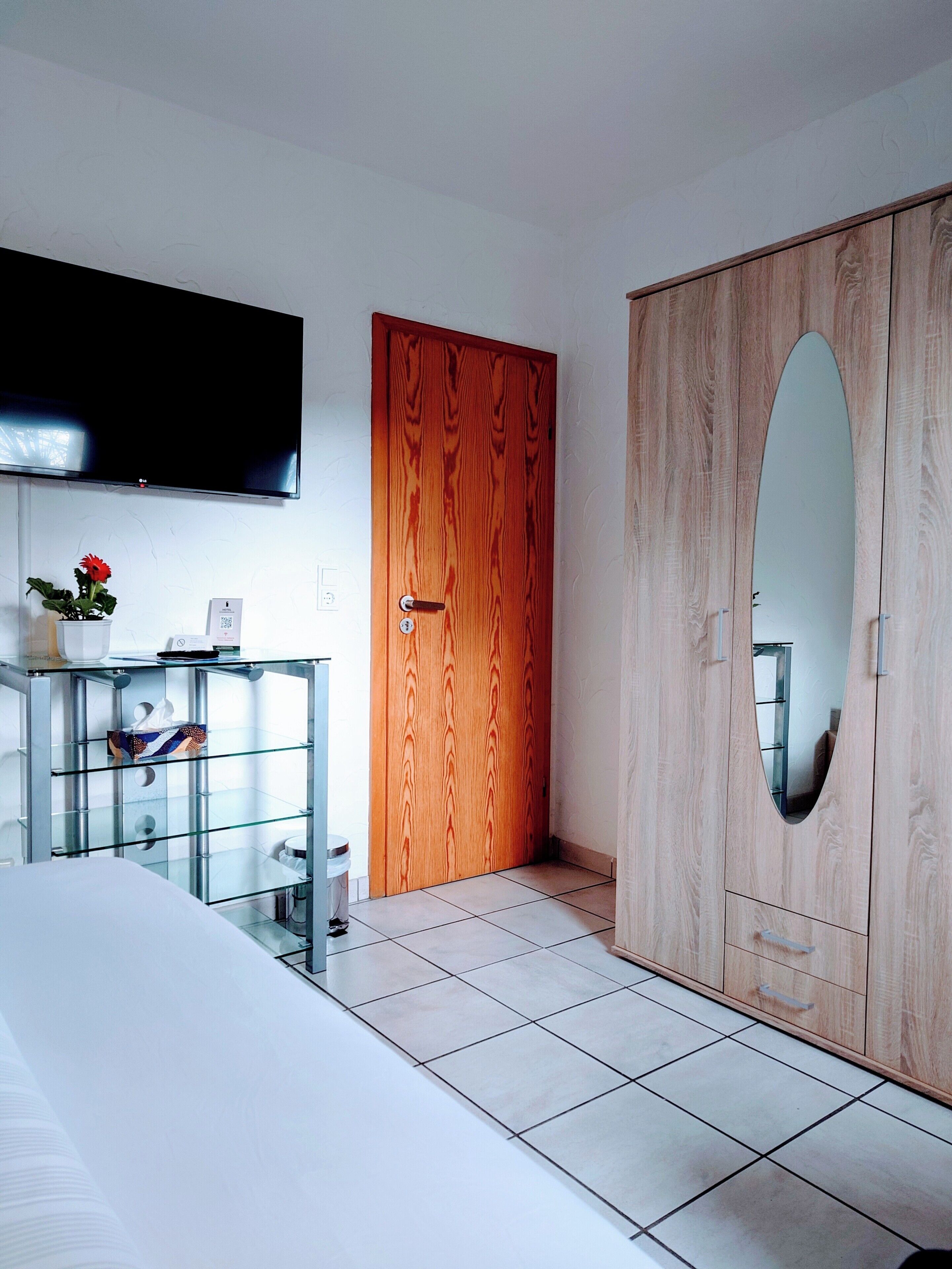 Quarto com cama, televisão na parede, guarda-roupa de madeira com espelho e aparador de vidro com planta.