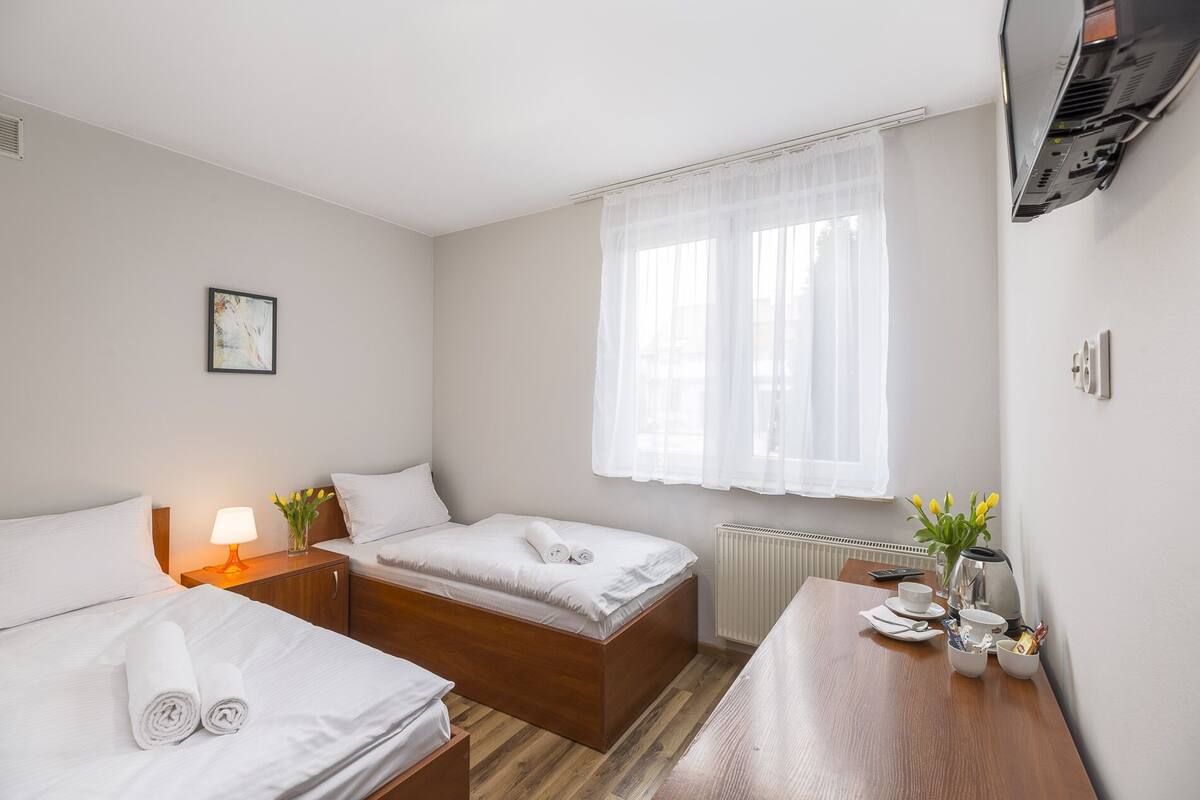 Quarto de hotel com duas camas, uma mesa de madeira com itens de café da manhã, um radiador e uma janela com cortinas leves.