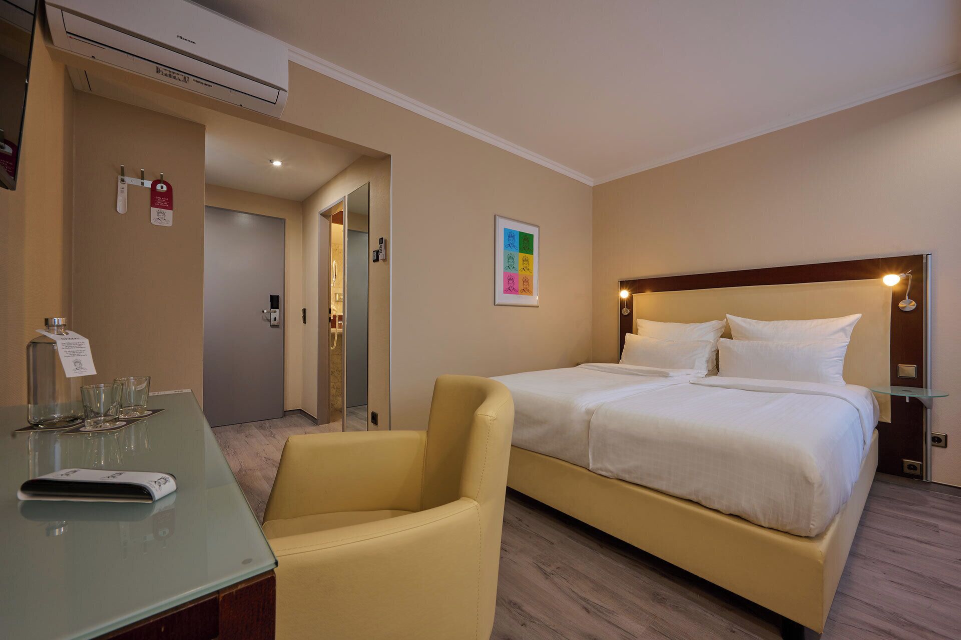 Quarto de hotel com cama, cadeira, mesa com copos e garrafa, e um quadro na parede.