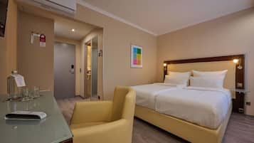 Quarto de hotel com cama, cadeira, mesa com copos e garrafa, e um quadro na parede.