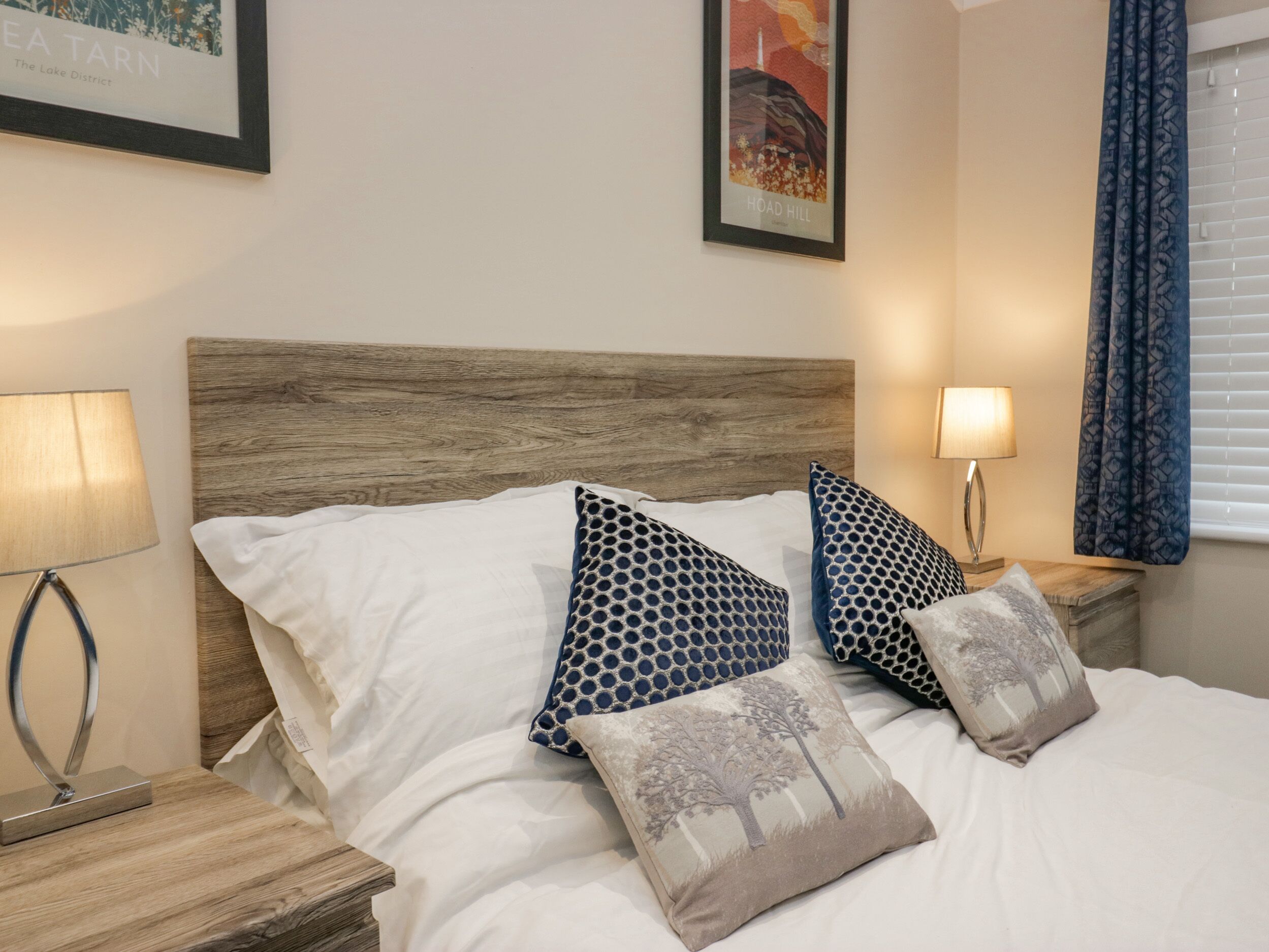 Quarto com cabeceira de madeira, roupa de cama branca, almofadas estampadas, abajures e um quadro na parede.