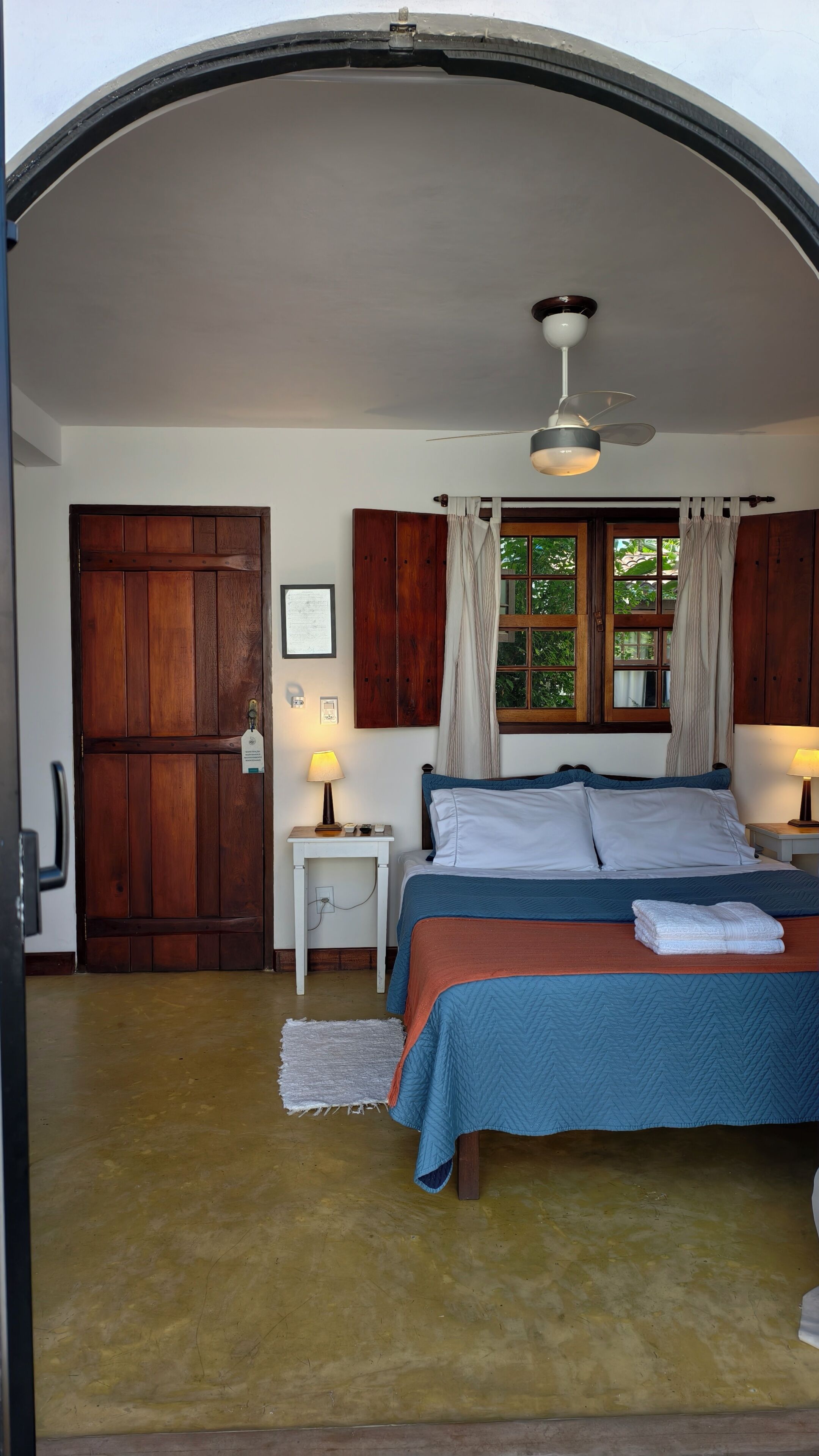 Um quarto com uma cama, duas mesas de cabeceira com abajures, um guarda-roupa de madeira, uma janela com cortinas e um ventilador de teto.
