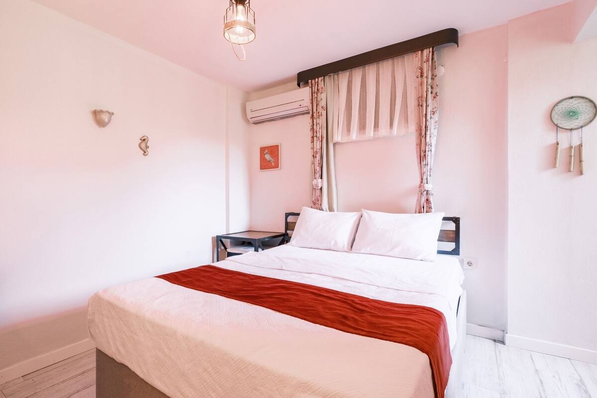 Um quarto com cama, mesa de cabeceira, ar-condicionado fixado na parede, um detalhe decorativo na parede e luminária de teto.