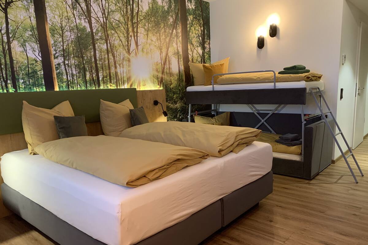 Quarto com uma cama grande, uma beliche e um mural na parede com tema de floresta.