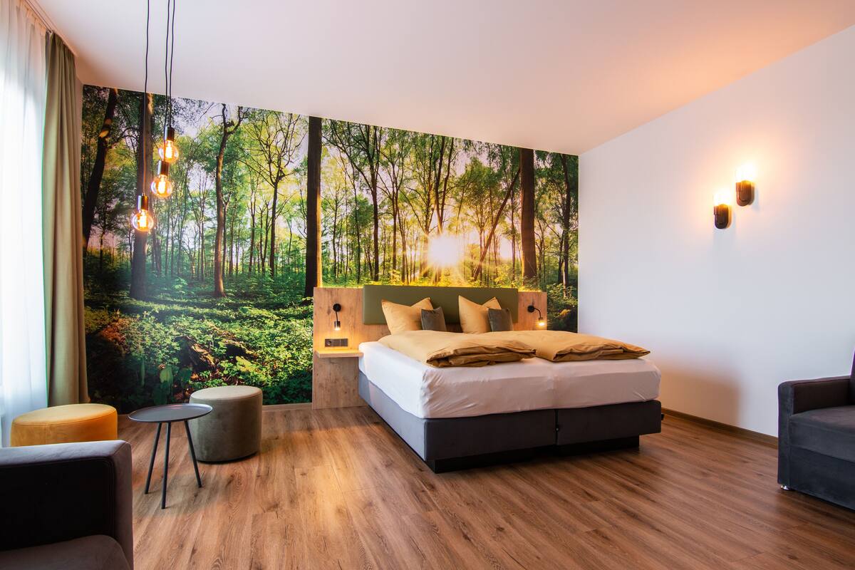 Quarto moderno com uma cama grande, mesa de cabeceira, mesa redonda e um sofá. Um mural com tema de floresta está na parede atrás da cama.