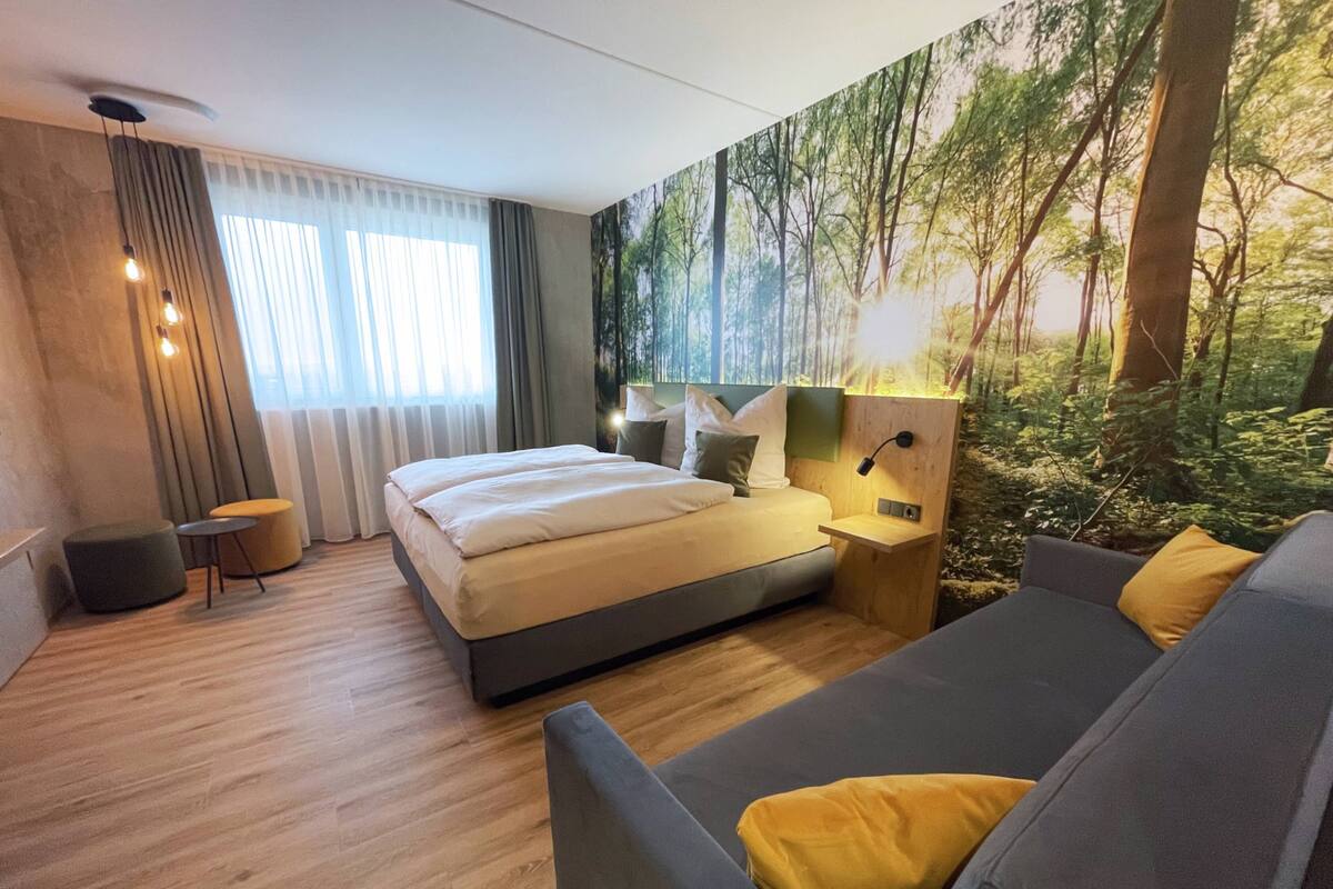 Quarto moderno com uma cama grande, um espaço de estar com sofá, uma mesinha redonda e um mural na parede com tema de floresta.