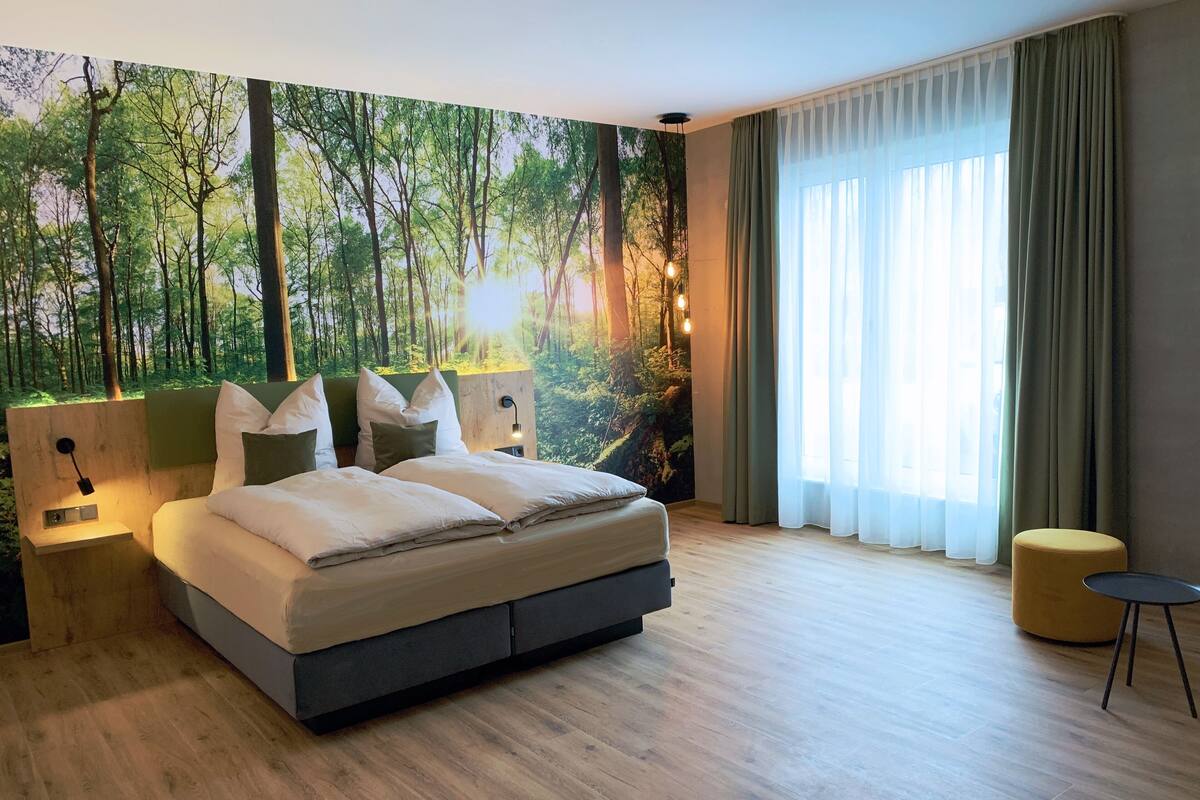 Quarto moderno com uma cama grande, uma mesinha de cabeceira, uma pequena mesa redonda e um mural na parede com uma cena de floresta.