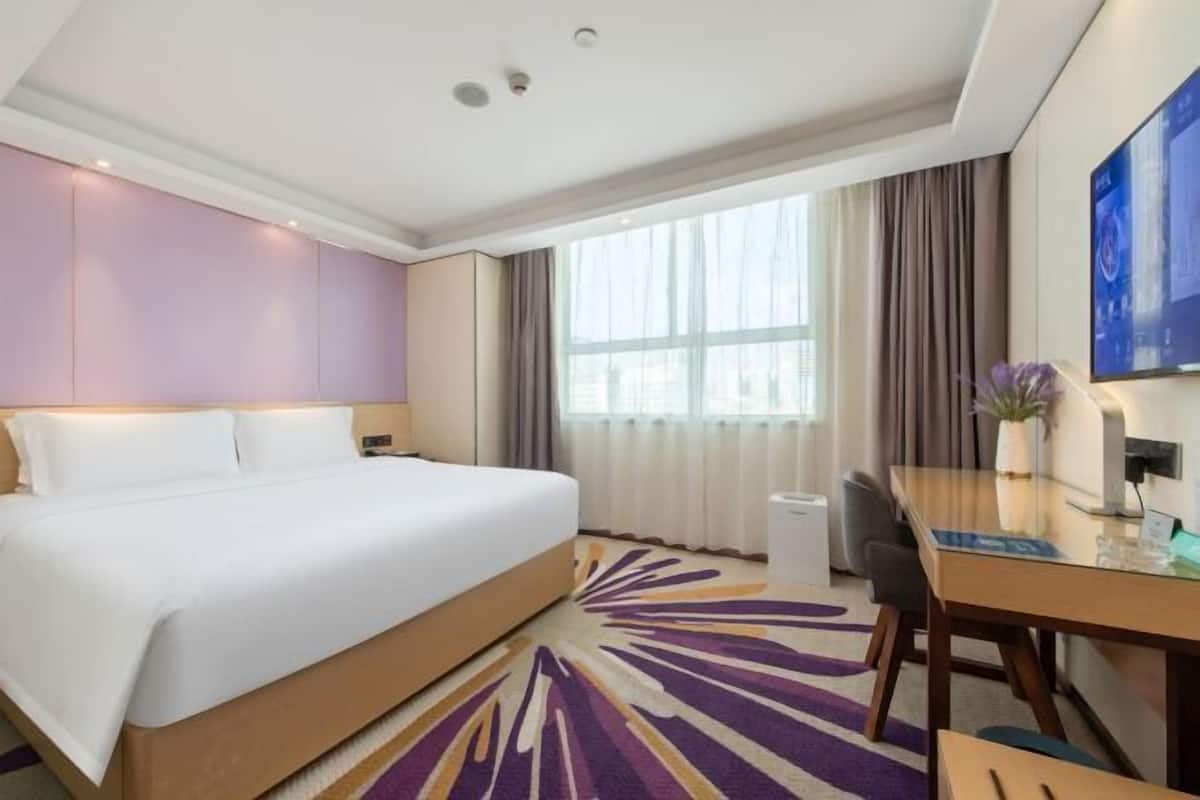 Quarto de hotel com uma cama grande, uma escrivaninha e uma cadeira. Há uma janela com cortinas, uma televisão de tela plana na parede e um tapete decorativo no chão.