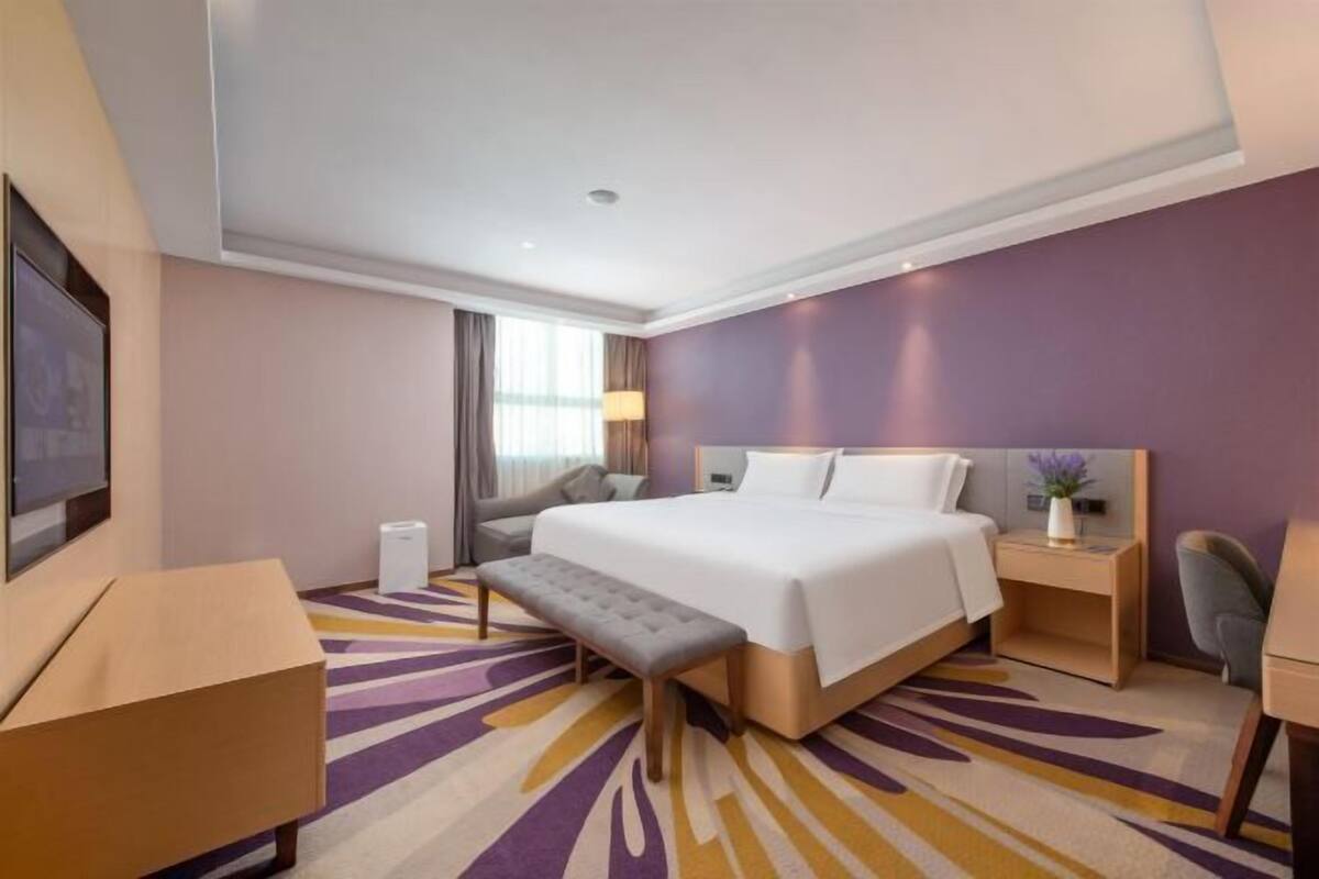 Quarto de hotel com cama, escrivaninha, cadeira, banqueta, mesa de cabeceira, abajur e janela.
