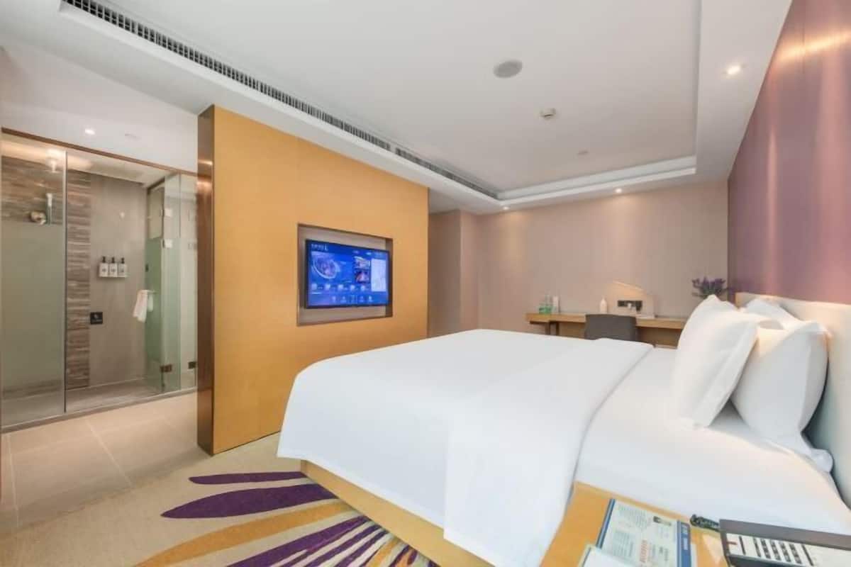 Quarto de hotel moderno com uma cama grande, uma tela digital na parede e área de chuveiro em vidro.