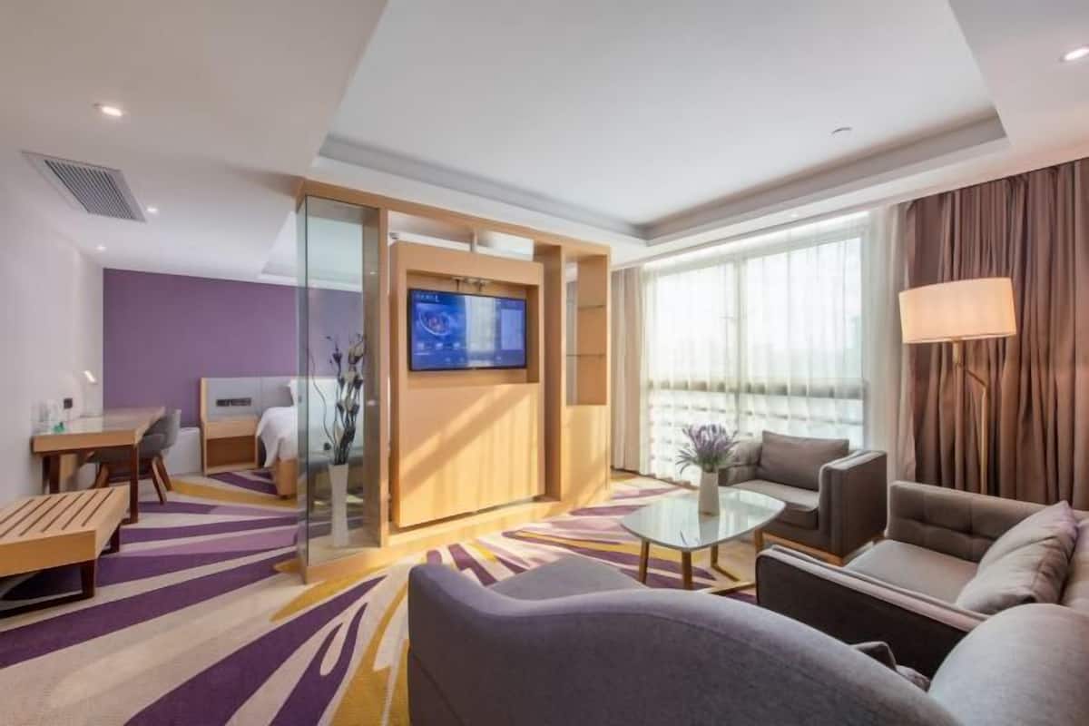 Quarto de hotel com cama, secretária, televisão, sofá, mesa de centro e uma janela grande com cortinas.