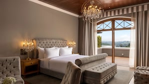 Quarto com cama grande, um candelabro, vista pela janela e um espaço de estar com uma poltrona e banquinho.