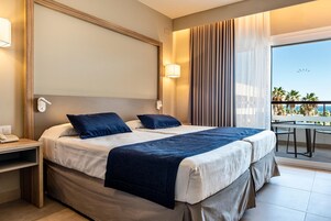 Quarto de hotel com duas camas, uma varanda com mesa e cadeiras, e vista para palmeiras e o mar.