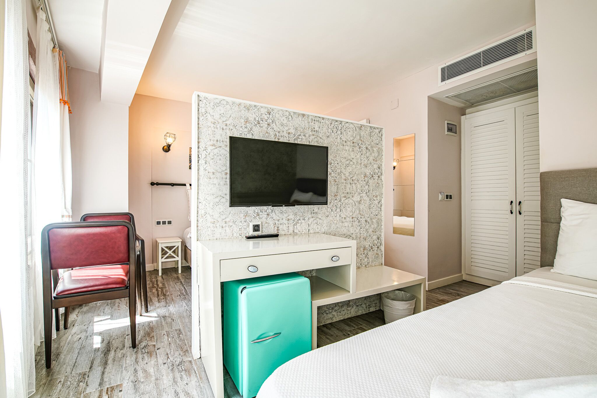 Quarto de hotel com cama, escrivaninha com televisão, pequeno frigorífico, cadeira e guarda-roupa.