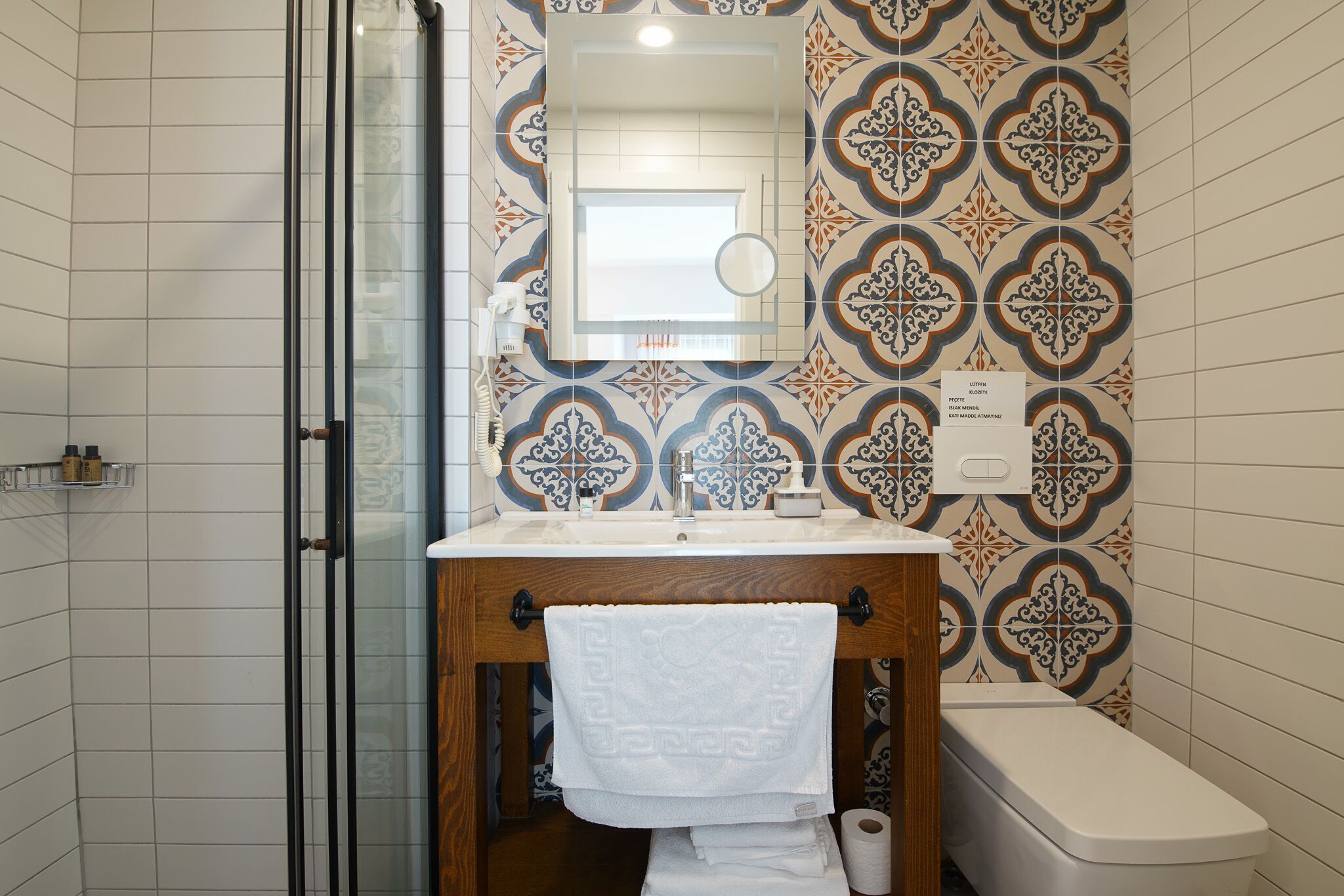 Casa de banho com chuveiro, pia com espelho, azulejos estampados e sanita.