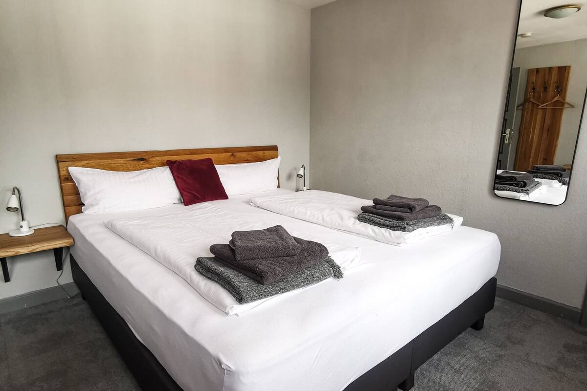 Cama bem arrumada com lençóis brancos e duas toalhas dobradas, cabeceira de madeira, mesa de cabeceira com abajur e um espelho grande na parede.