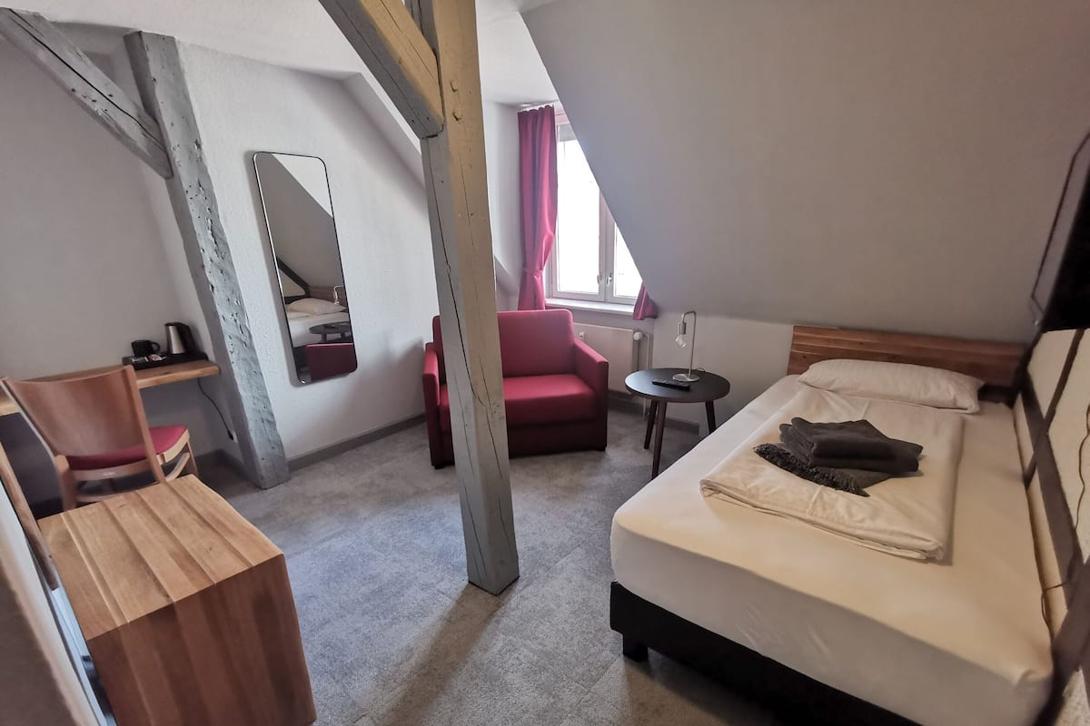 Quarto de hotel pequeno com uma cama, uma poltrona vermelha, uma cadeira de madeira, uma mesinha e um espelho na parede.