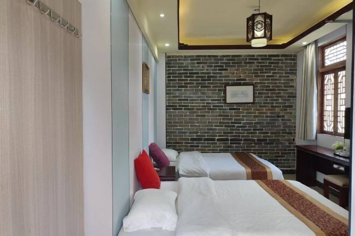 Quarto de hotel com duas camas, parede de tijolos, escrivaninha e janela com cortinas.