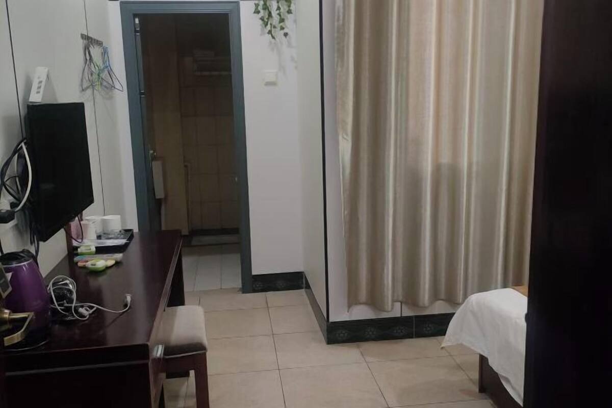 Quarto de hotel com uma cama, uma secretária com um computador e monitor, uma cadeira, uma mesinha com chávenas e uma planta, e uma cortina.