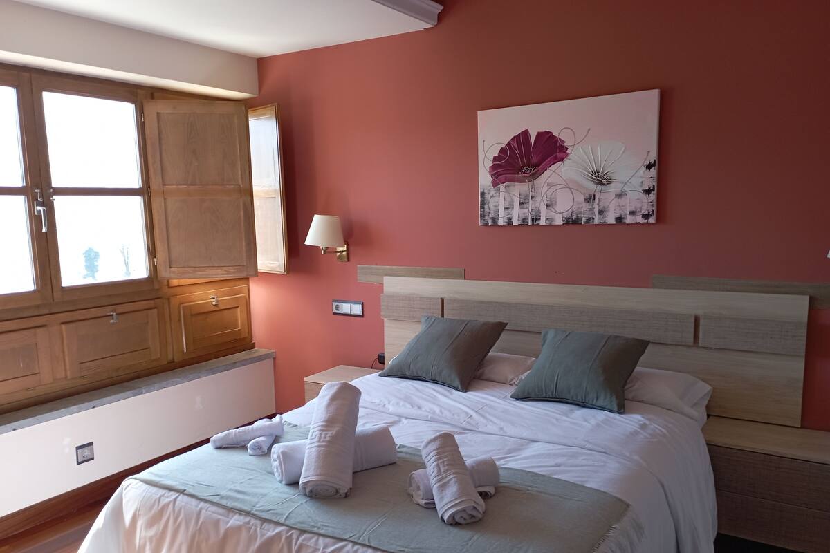 Cama bem arrumada com travesseiros e toalhas dobradas, cabeceira de madeira, luminária na parede e janela com persianas de madeira.
