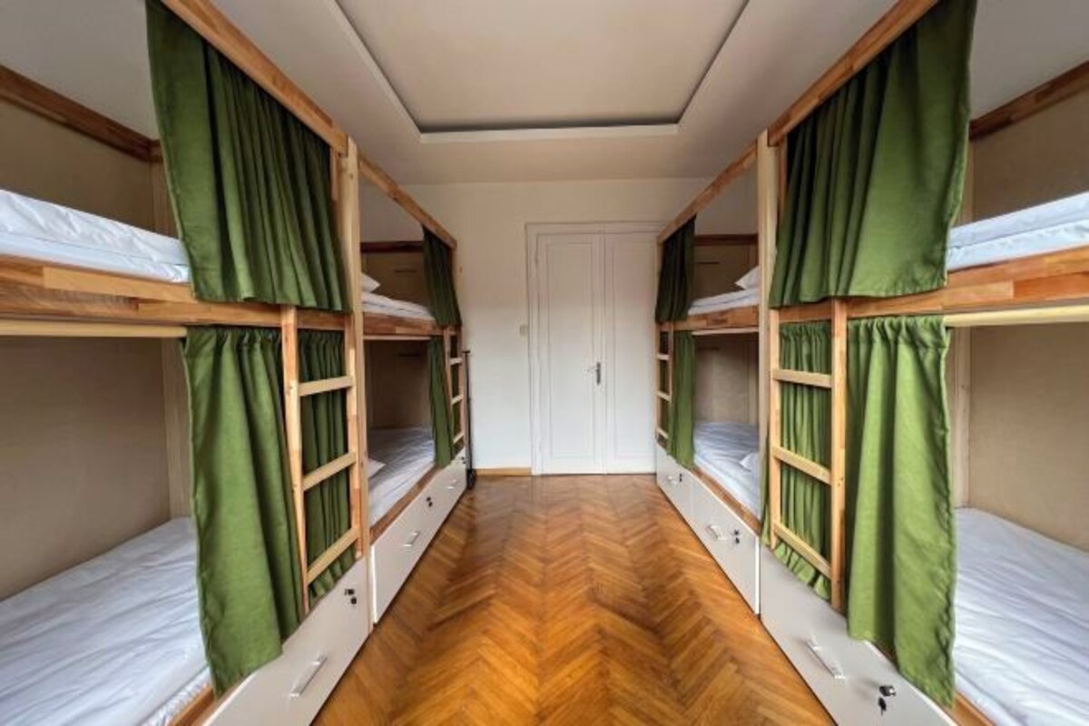 Quarto com quatro beliches dispostos em um corredor, cada um com estrutura de madeira e cortinas verdes.