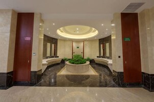 Um saguão de hotel com uma área de estar circular central, uma mesa de recepção e um espelho grande.