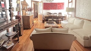 Uma sala de estar com um sofá branco, piso de madeira, um espaço de jantar com cadeiras e uma mesa, e um espelho grande à esquerda.