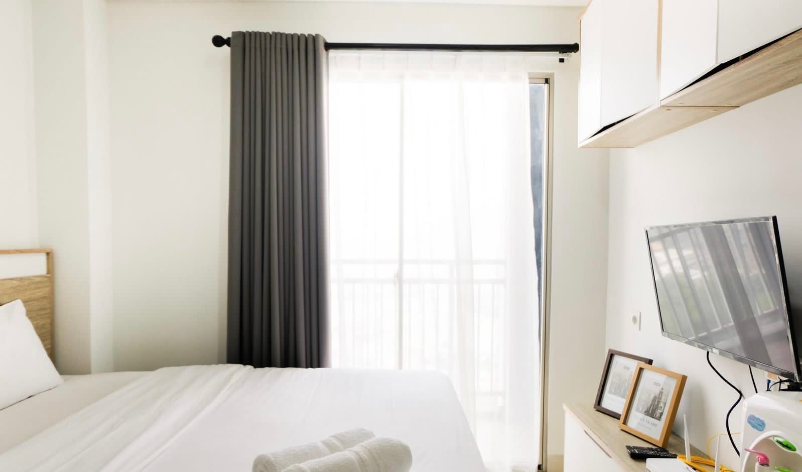 Quarto de hotel com uma cama, televisão, secretária com computador e janela com cortinas.