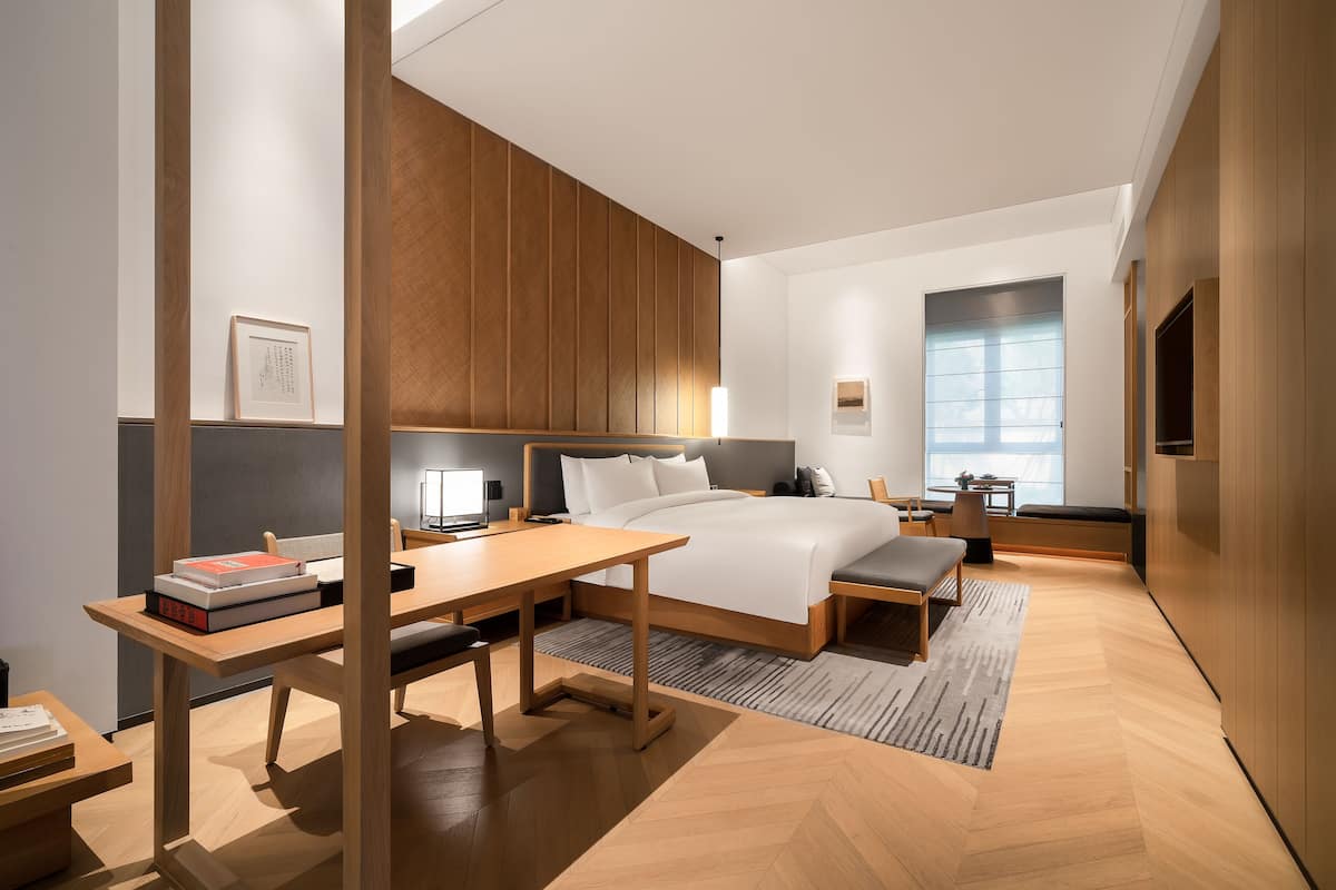 Quarto de hotel moderno com cabeceira de madeira, uma cama grande, escrivaninha com cadeira e janela com persianas.