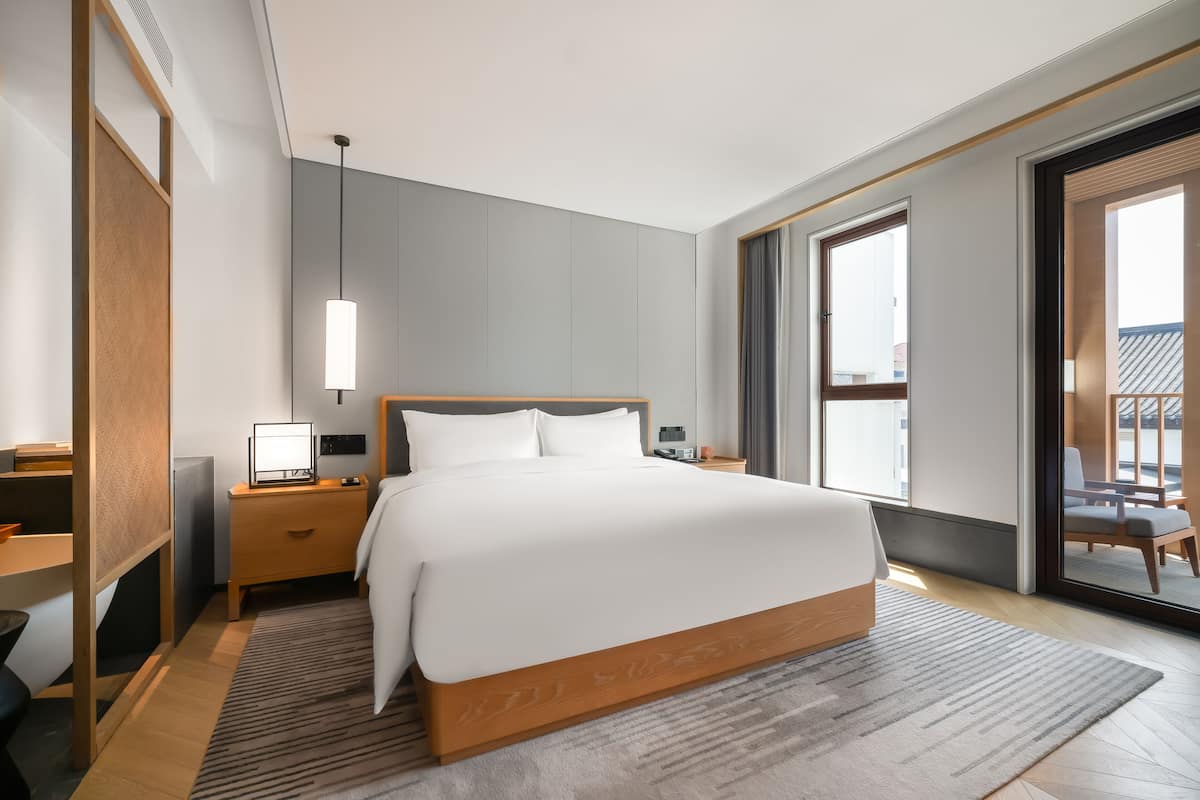 Quarto de hotel moderno com uma cama grande, cabeceira de madeira, mesa de cabeceira com abajur, janela ampla e vista para o exterior.