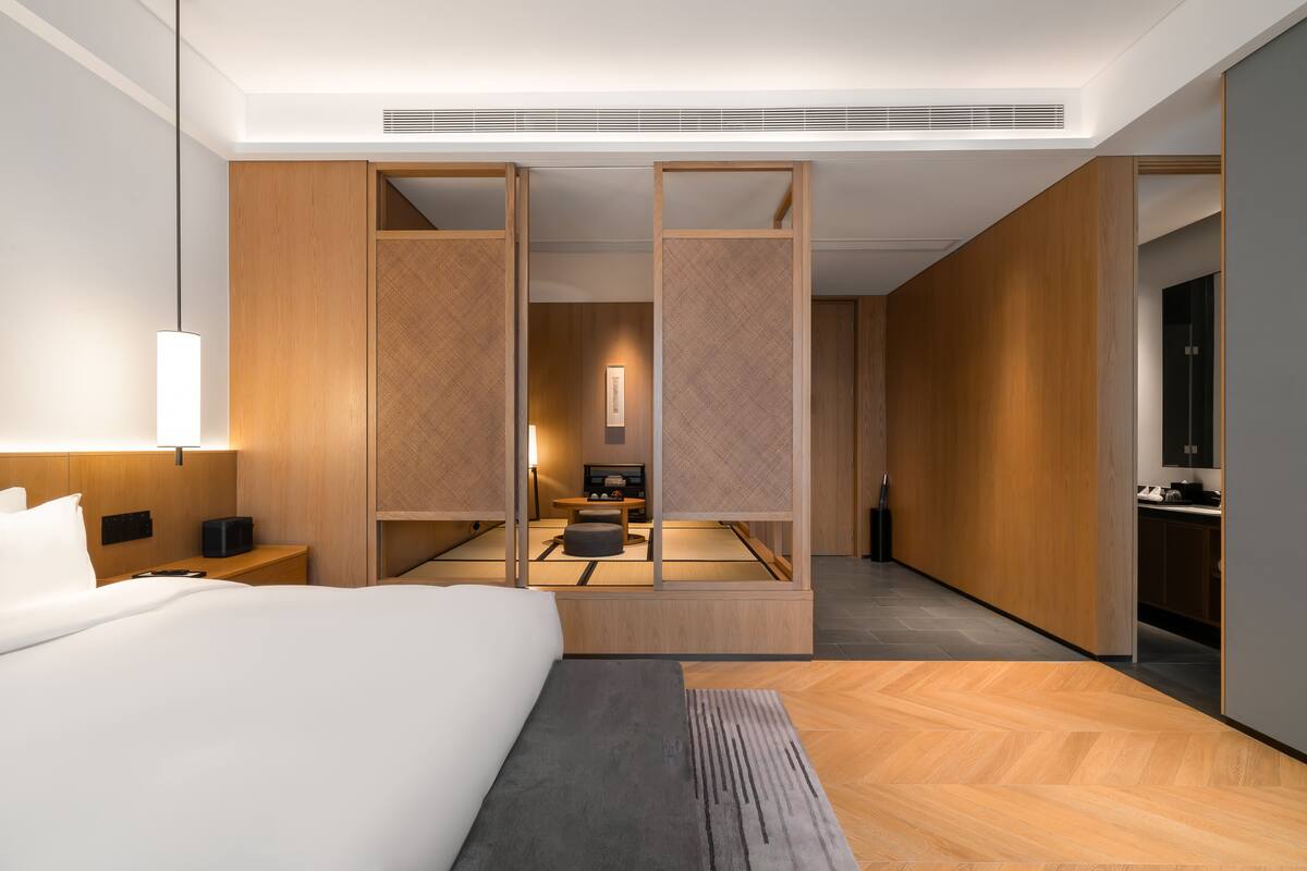 Quarto de hotel moderno com uma cama grande, uma mesa de trabalho, área de estar e vista através de um separador.