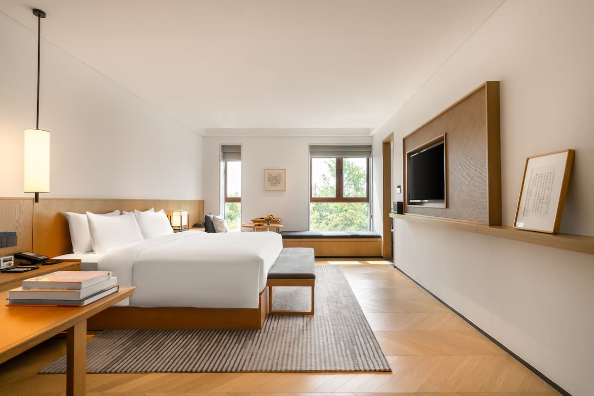 Quarto de hotel moderno com uma cama grande, uma escrivaninha com livros, um banco, uma TV na parede e uma janela com vista para o verde.