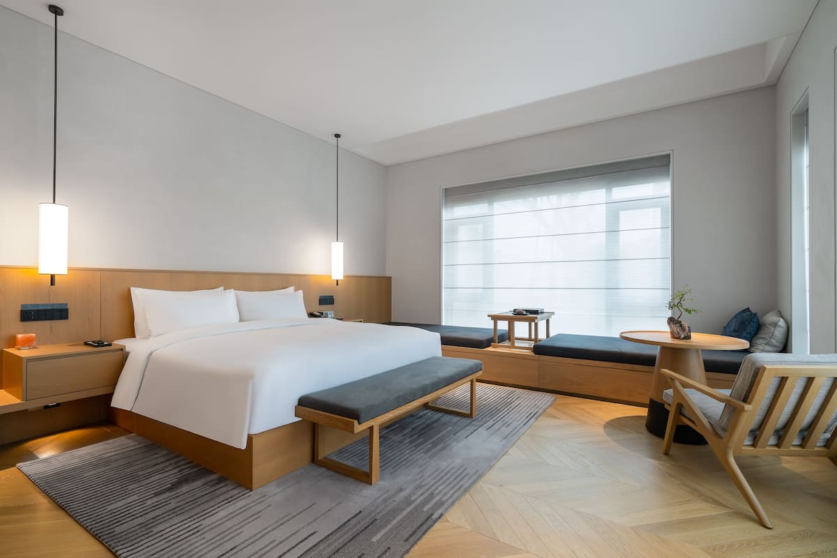 Quarto de hotel moderno com uma cama grande, mesas de cabeceira, um banco, uma mesa redonda e uma janela com persianas.